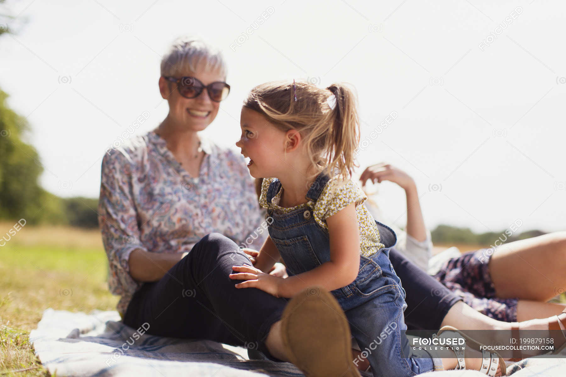 Grandmother And Granddaughter Laughing On Blanket In Sunny Field