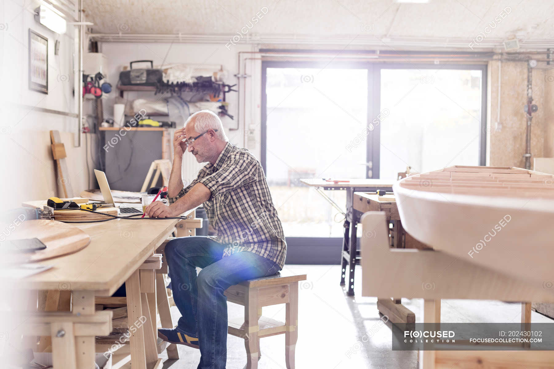 Male carpenter working at laptop on workbench near wood boat in ... Male carpenter working at laptop on workbench near wood boat in ...