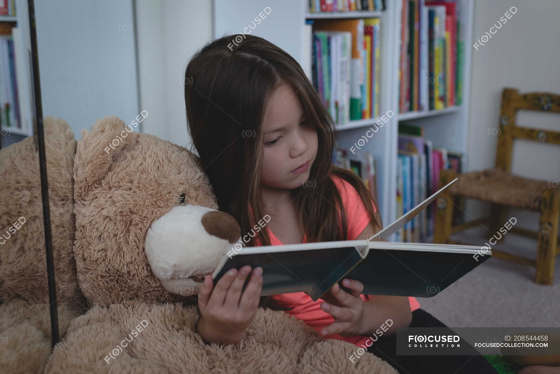 Elementary age child with teddy bear reading book in living room at ...