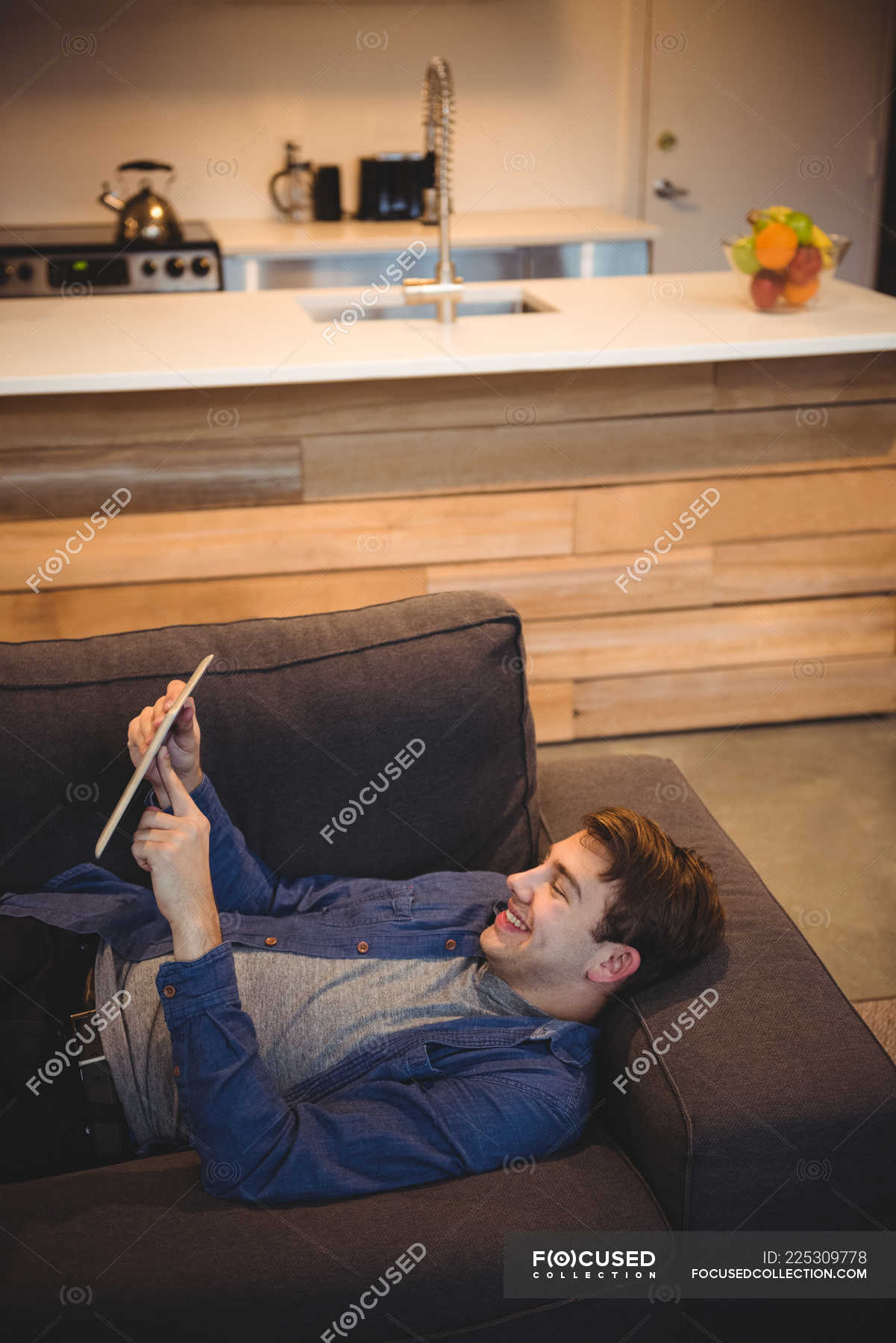 Smiling man lying on sofa using digital tablet in living room at home ...