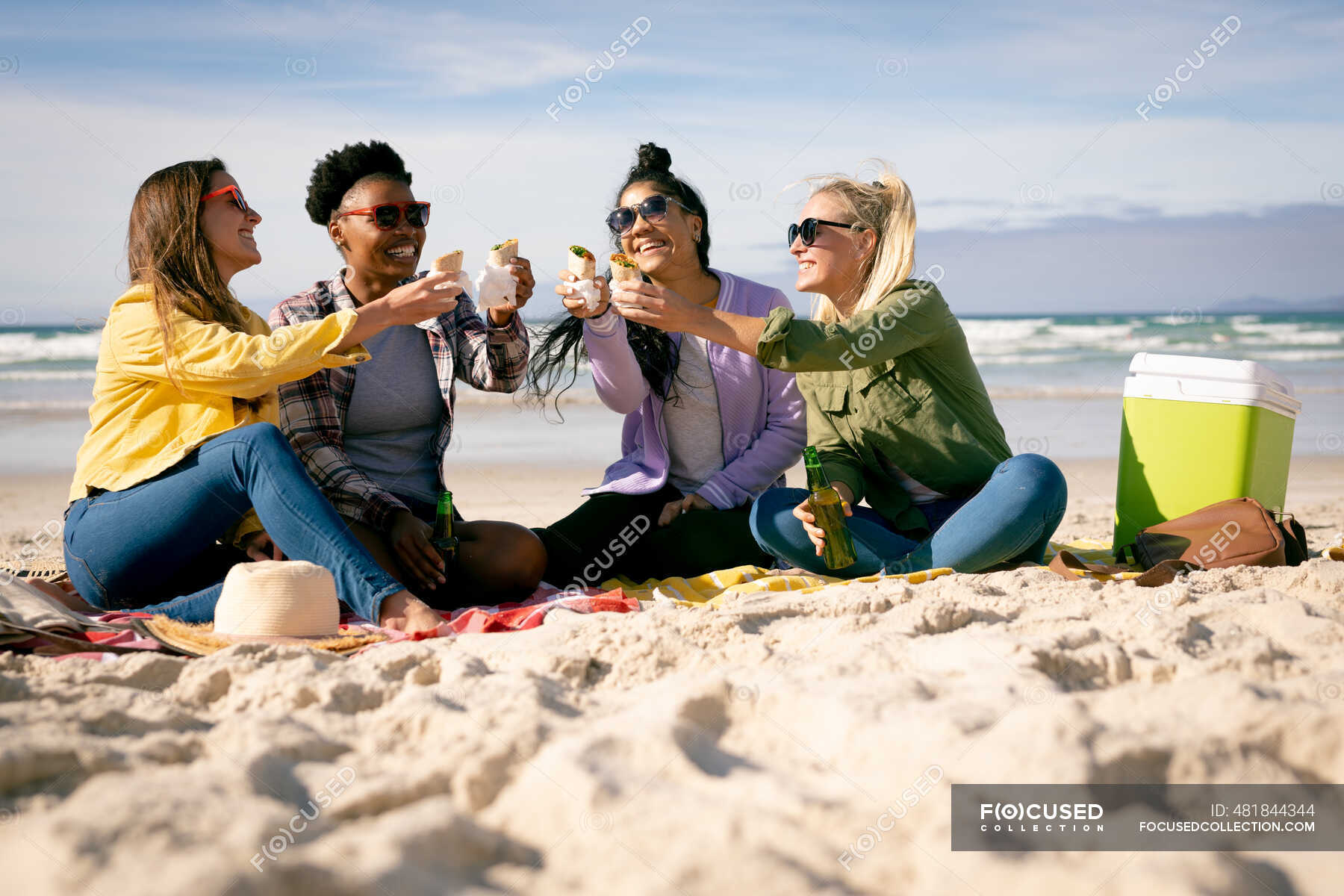 Happy group of diverse female friends having fun, siting on beach holding food laughing. holiday ...