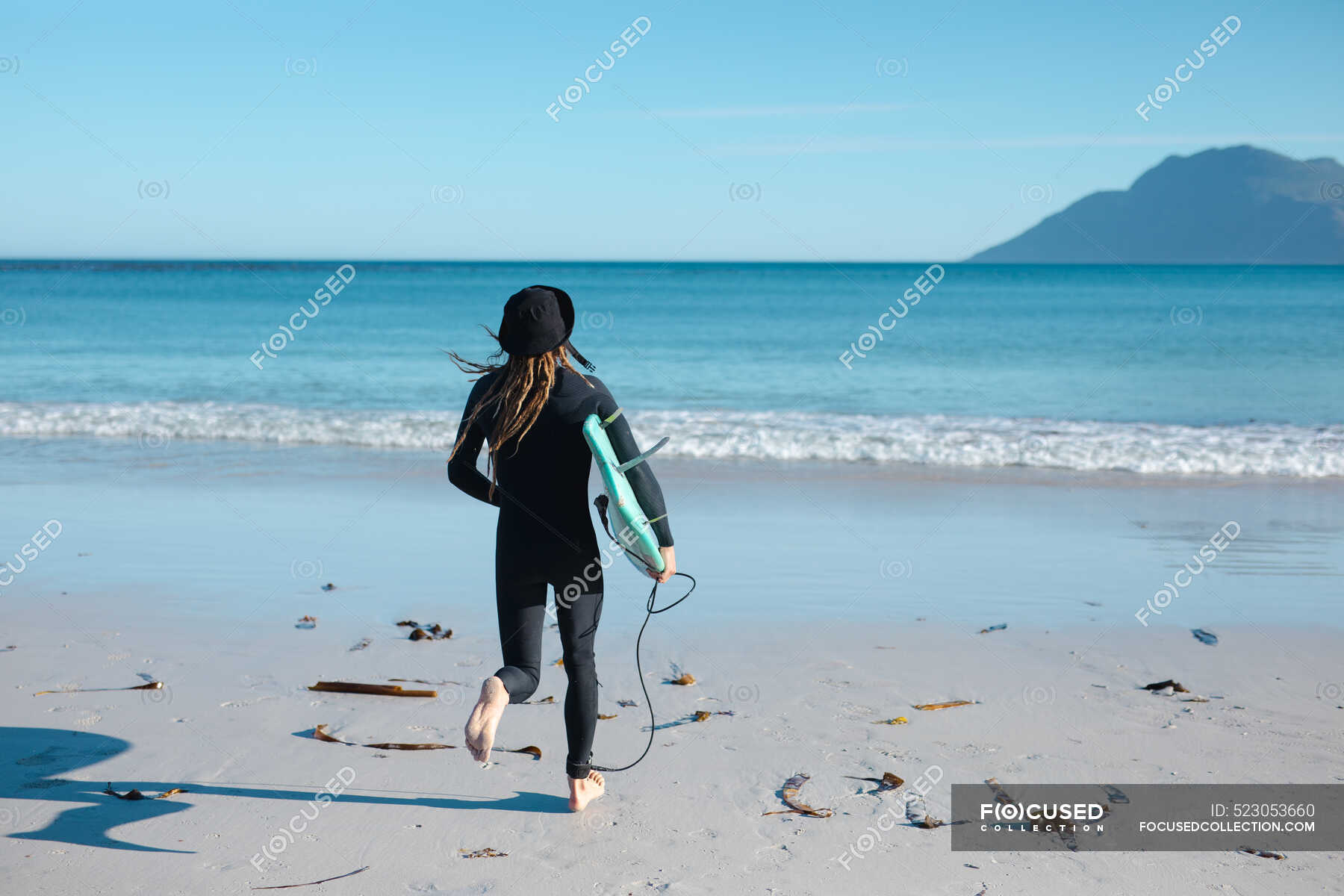 Rear view of male surfer running with surfboard on shore towards sea ...