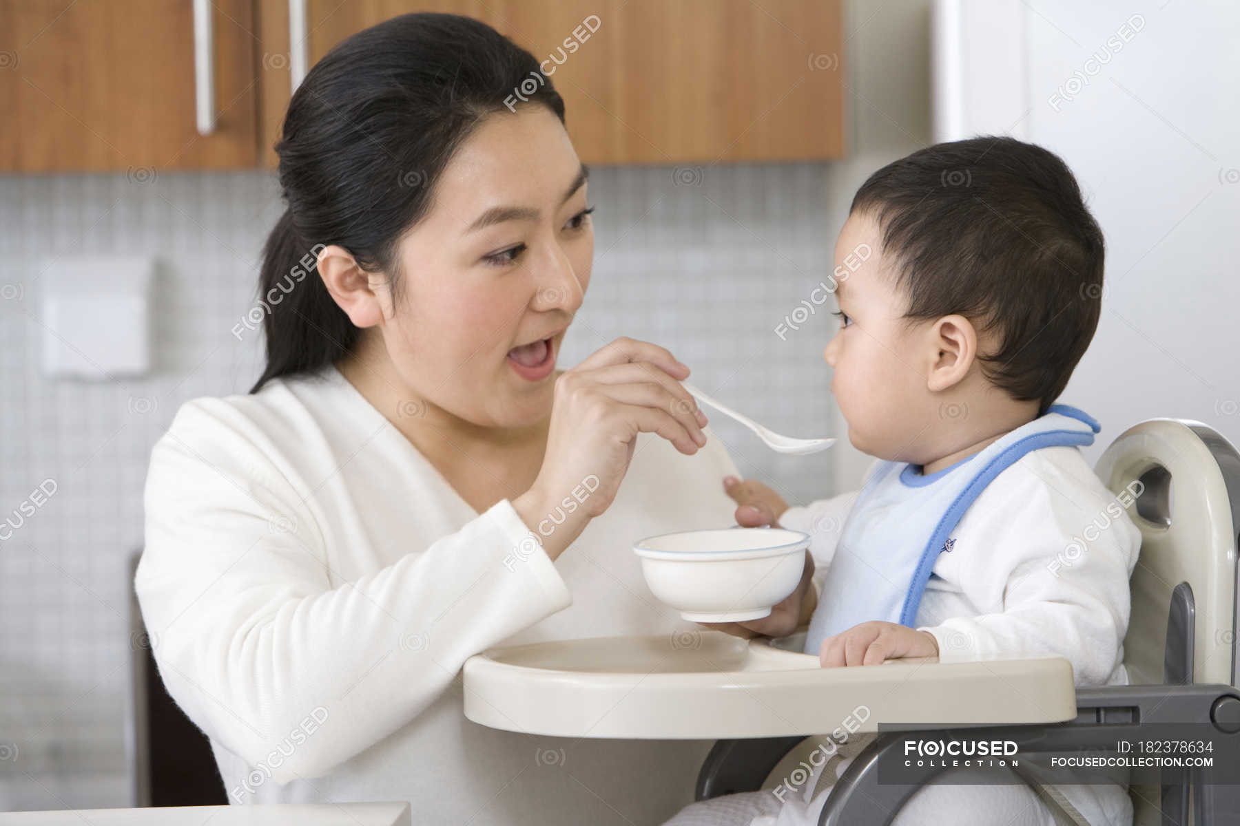 Chinese woman feeding baby son in high chair in kitchen — One Parent ...