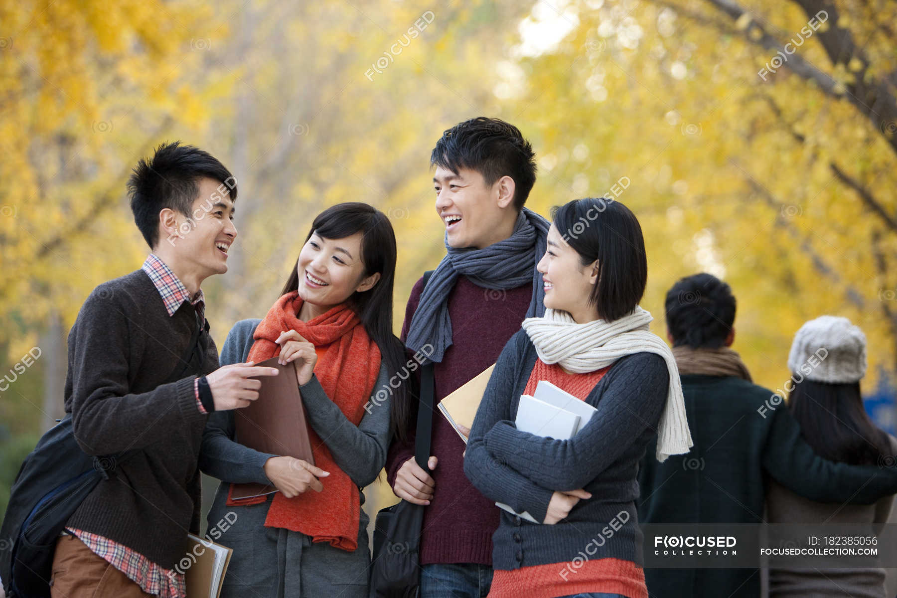 Chinese college students with books talking in campus park in autumn ...