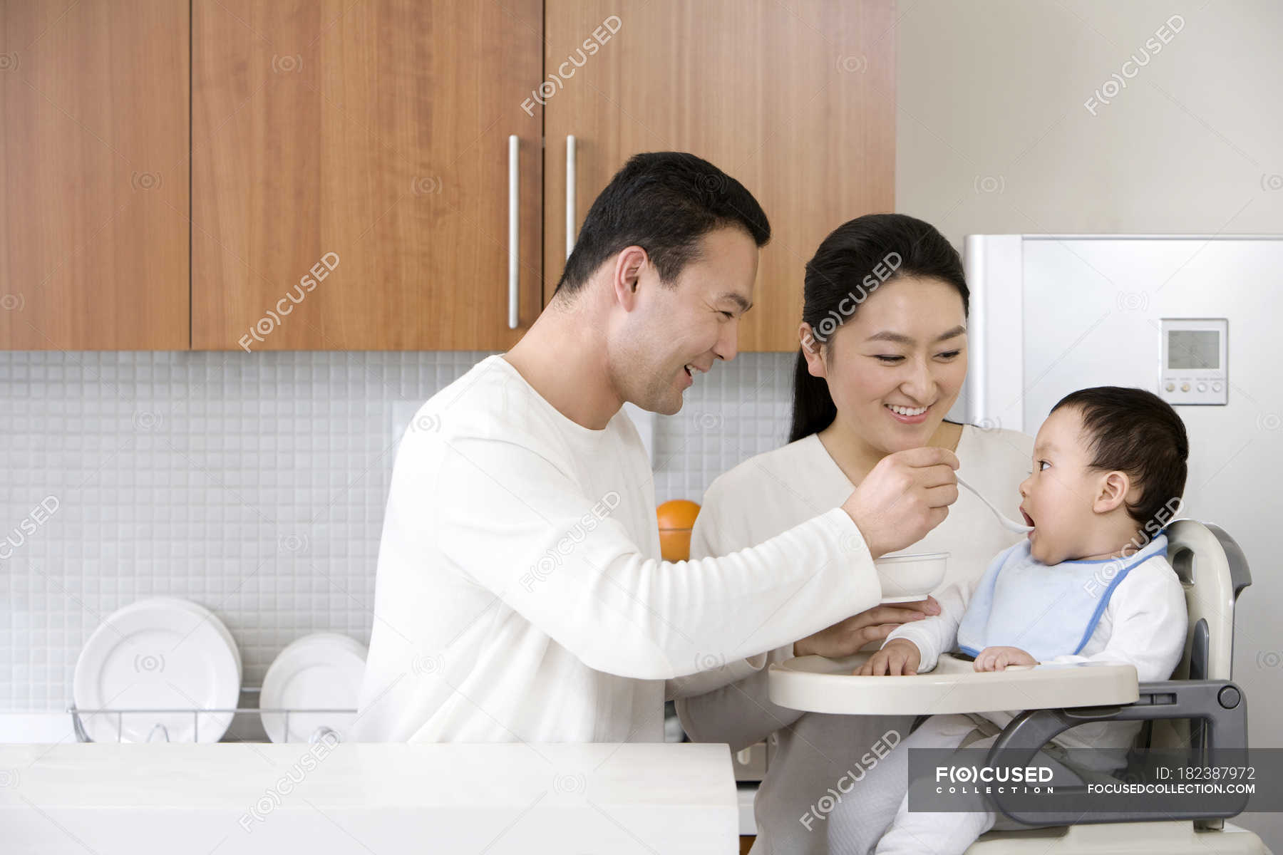 Chinese parents feeding baby boy in high chair — standing, young family ...
