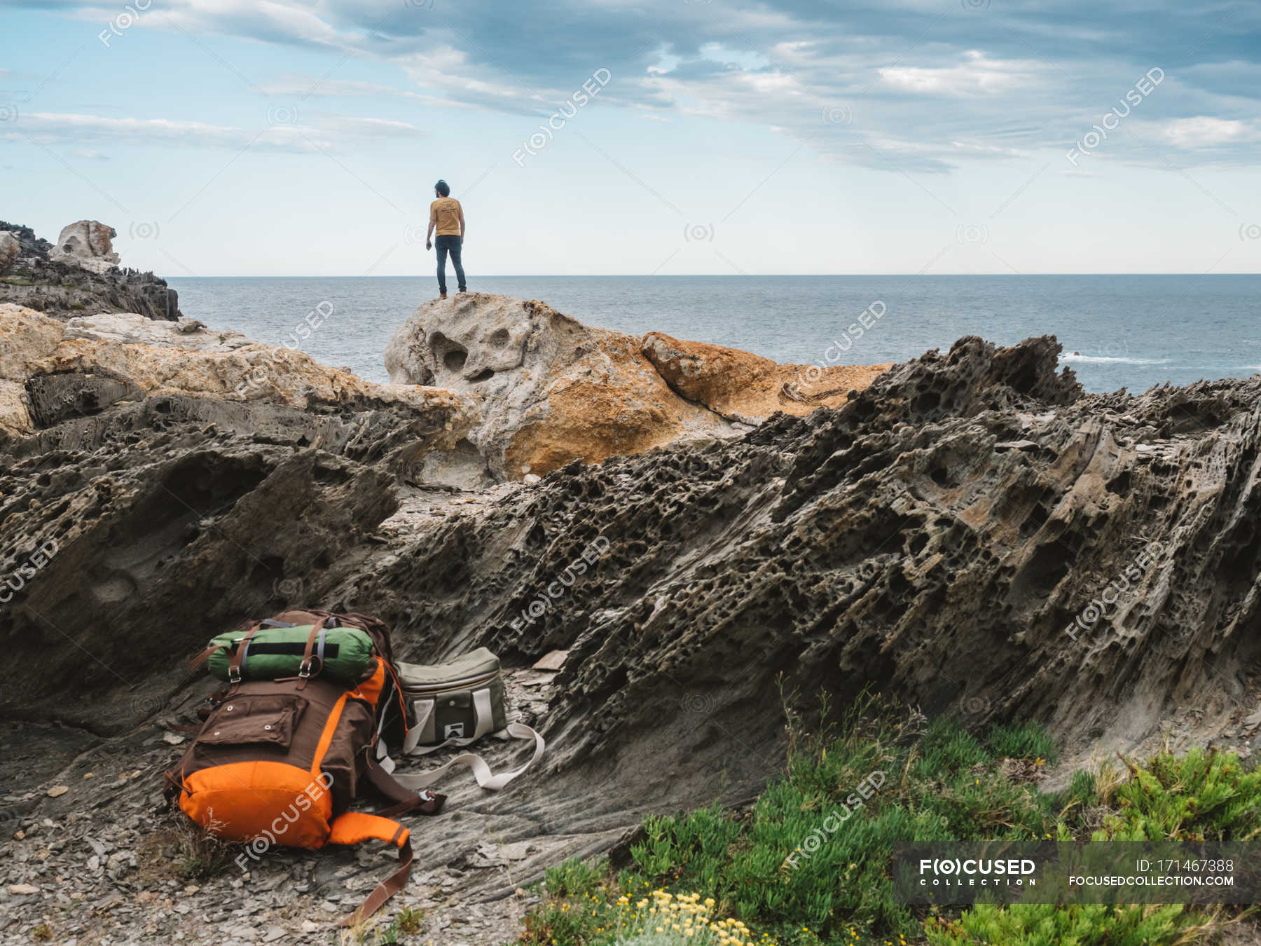 Travel backpack lying on rocks over traveler standing on rock cliffs and admiring oceanscape ...