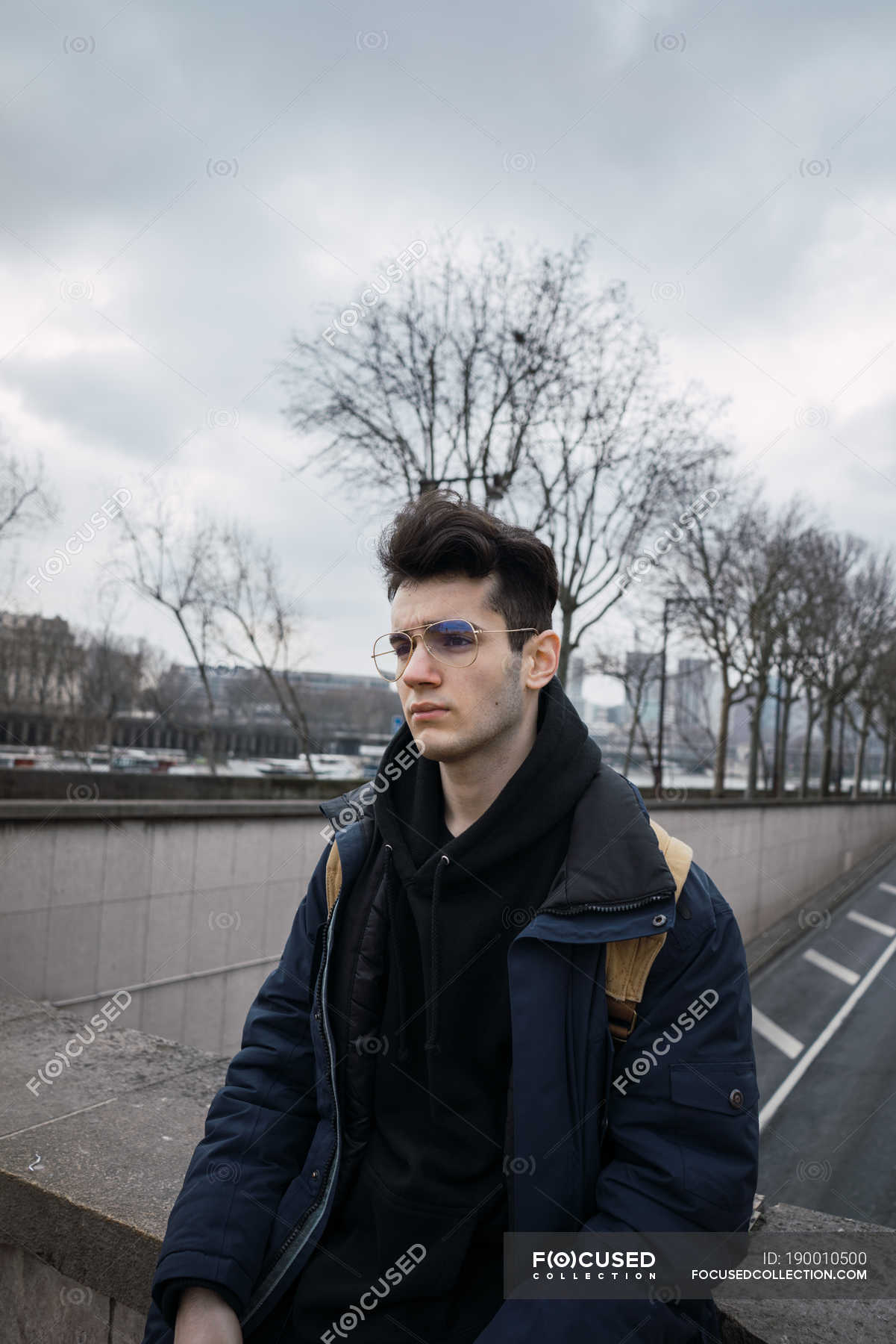 Thoughtful young man in glasses posing on background of asphalt road in ...
