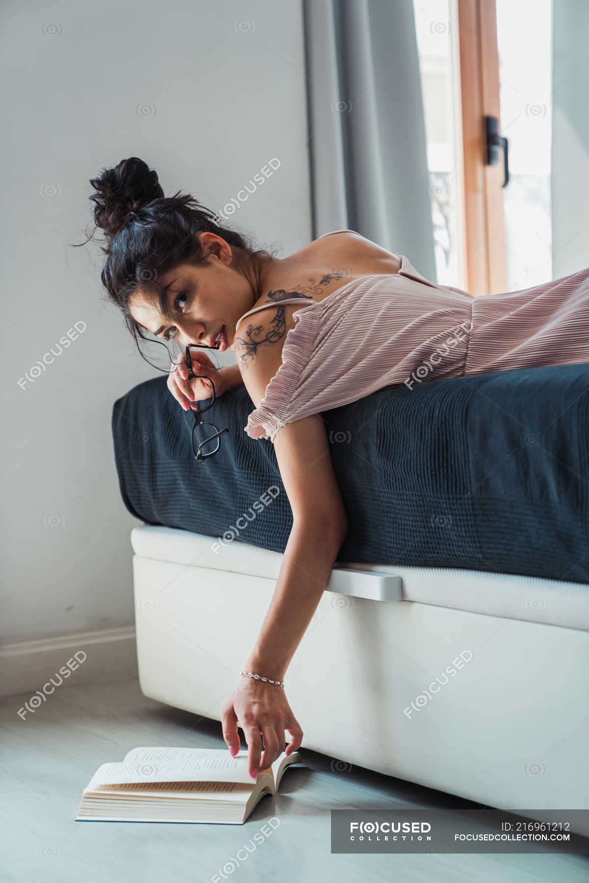 Flirty woman lying on bed with book on floor and biting eyeglasses ... Flirty woman lying on bed with book on floor and biting eyeglasses ...