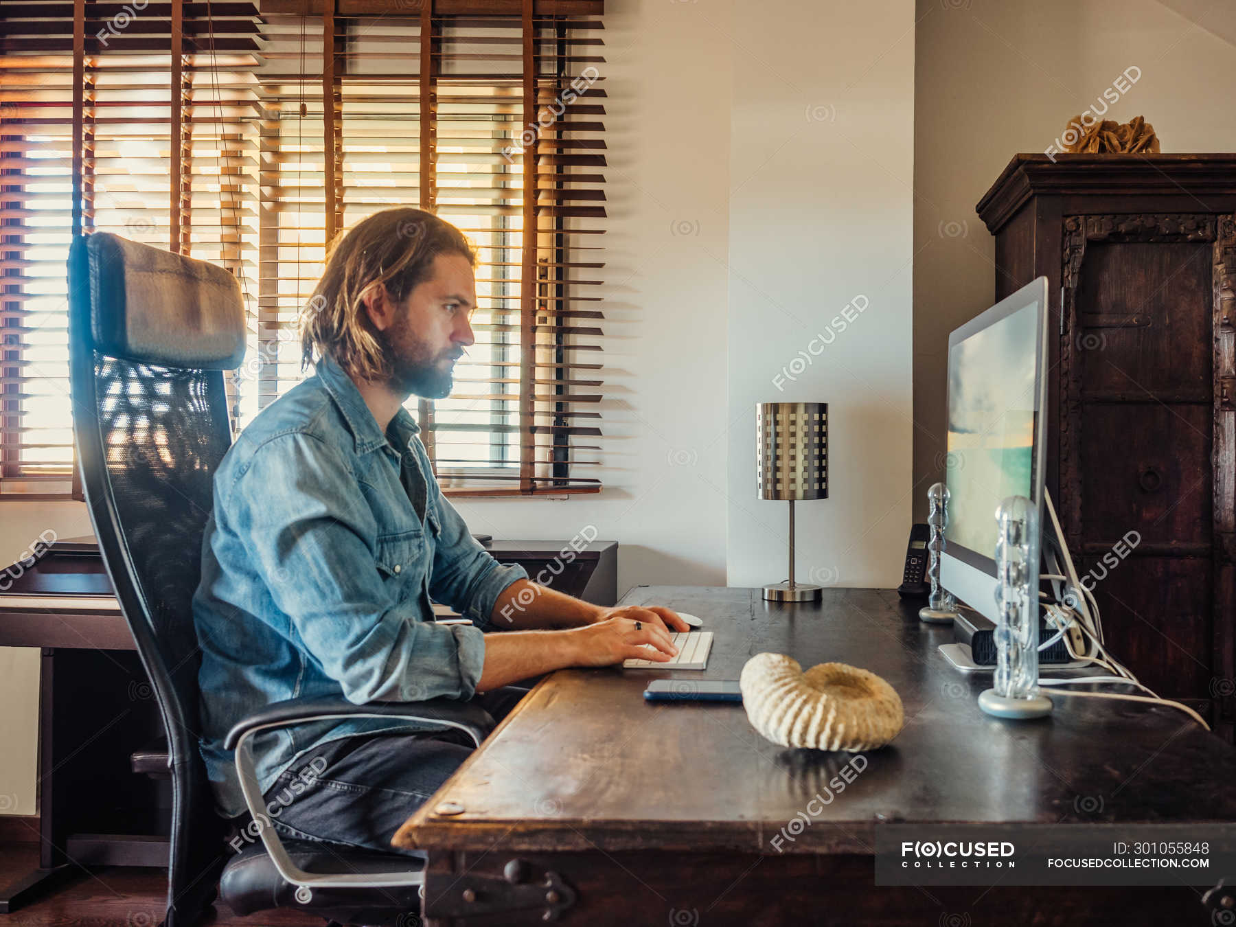Side view of serious man in denim shirt sitting in comfy armchair while ...