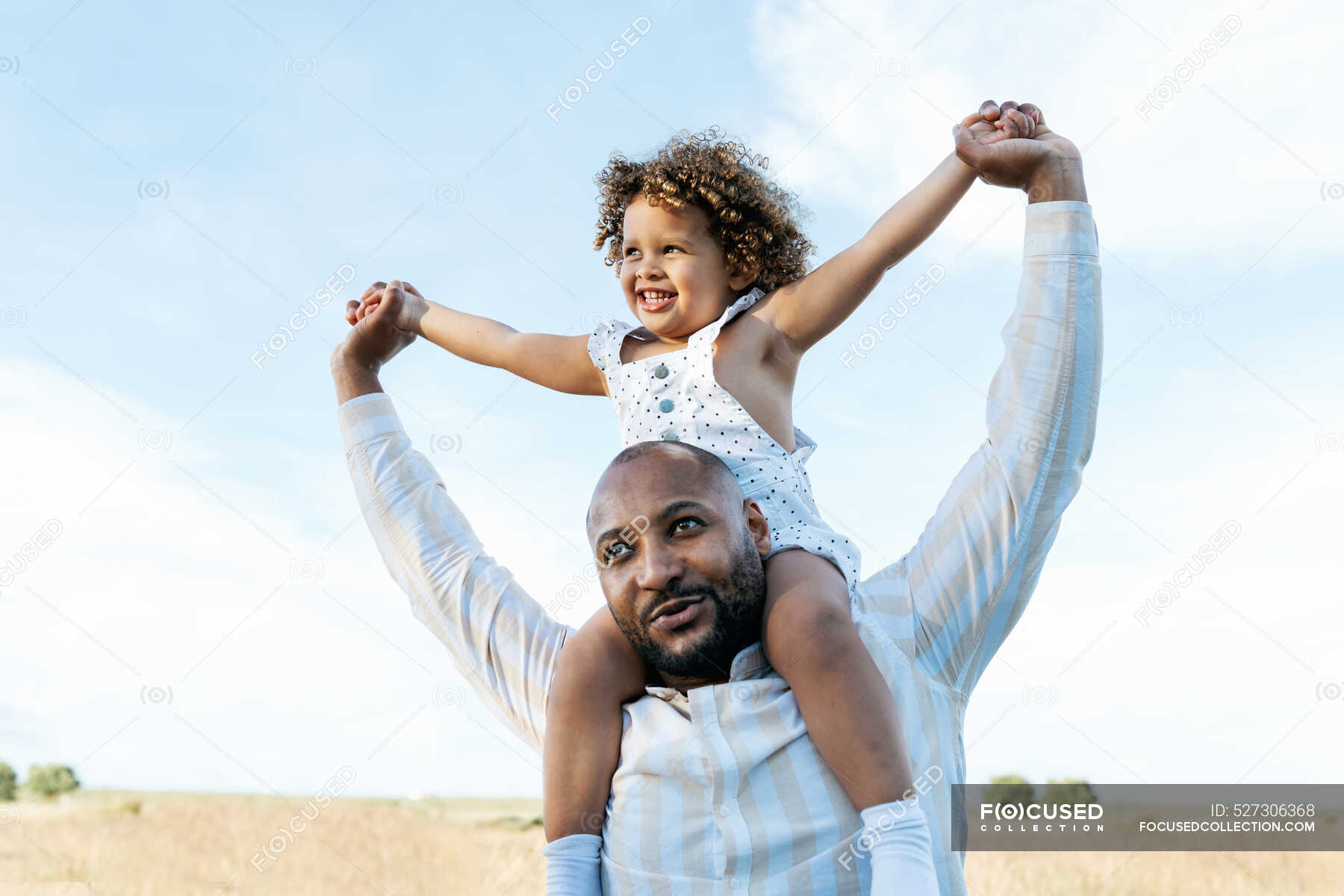 Cheerful African American father with cute little daughter on shoulders playing in field in ...