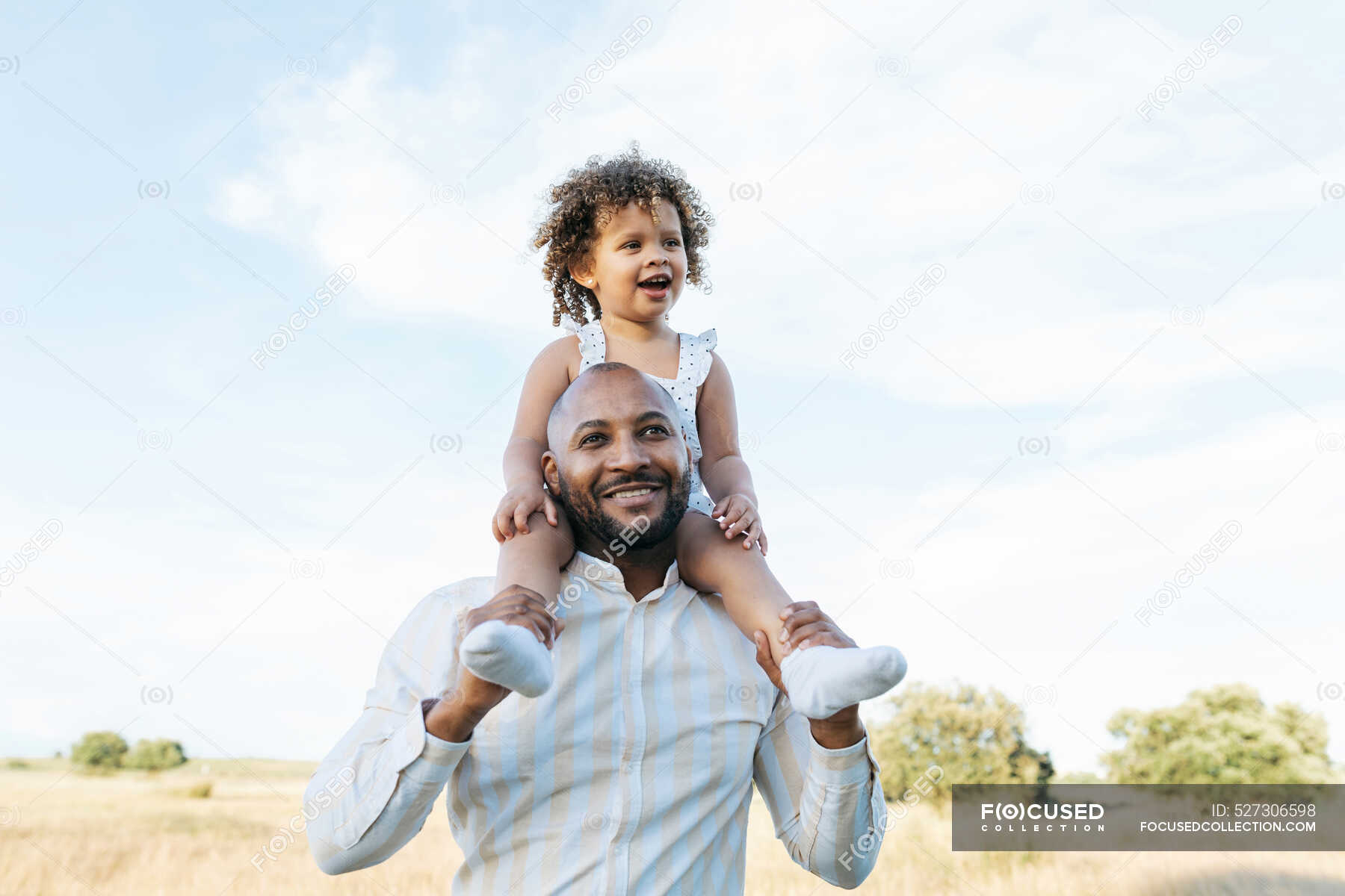 Cheerful African American father with cute little daughter on shoulders playing in field in ...